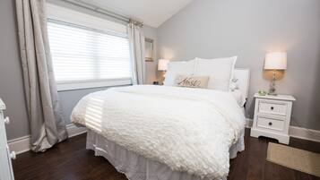 Quarto com uma cama grande, roupa de cama branca, mesa de cabeceira com abajur e janela com cortinas.