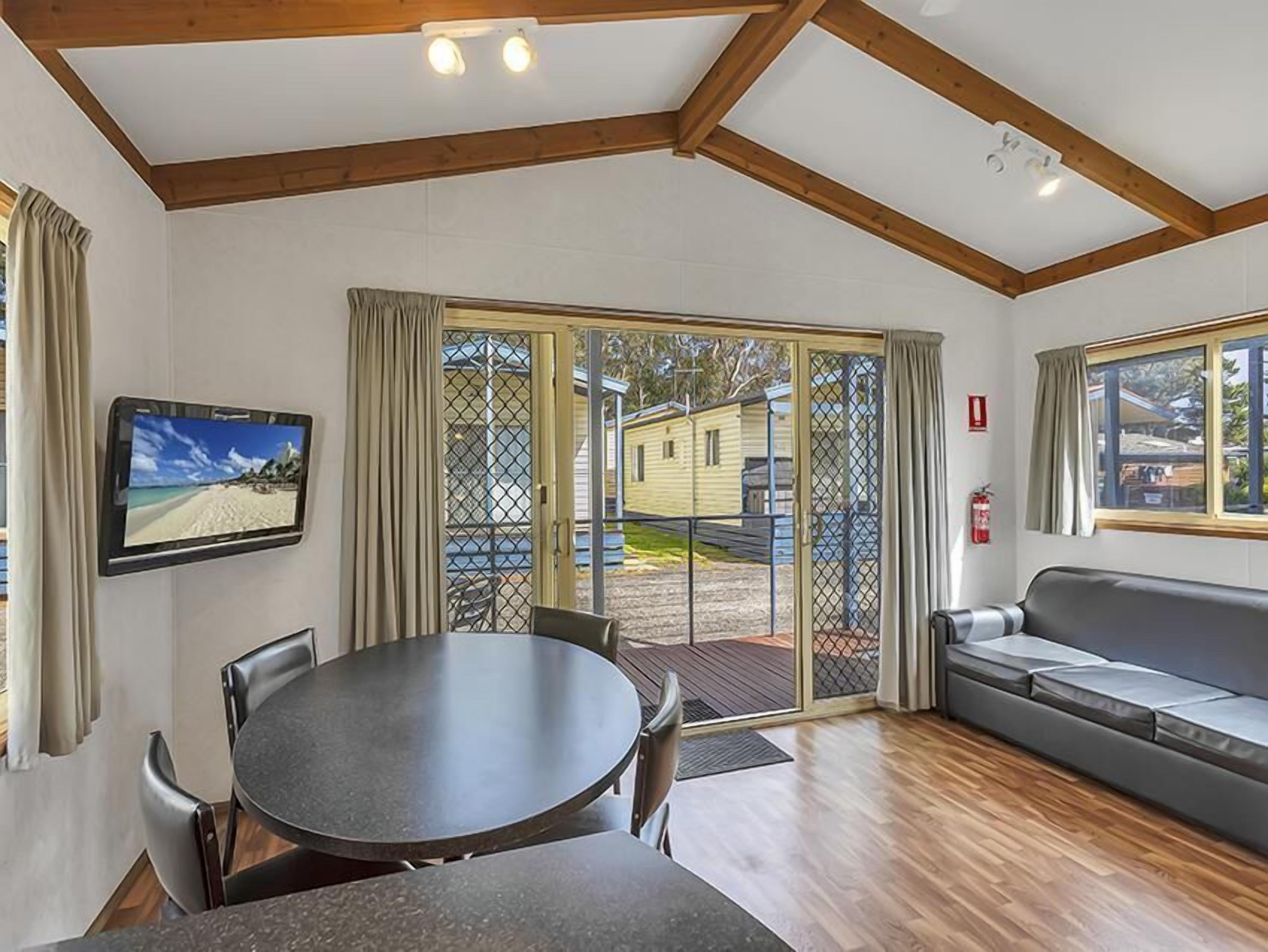 Quarto com mesa redonda, cadeiras, sofá e porta de correr de vidro que dá para área externa com vista para uma casa e árvores.