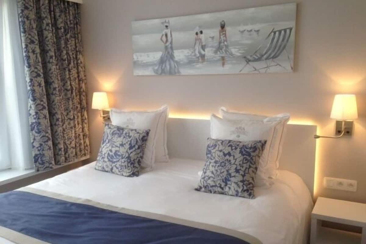 Quarto de hotel com uma cama grande, duas almofadas estampadas, um quadro sobre a cama e duas luminárias fixadas na parede.