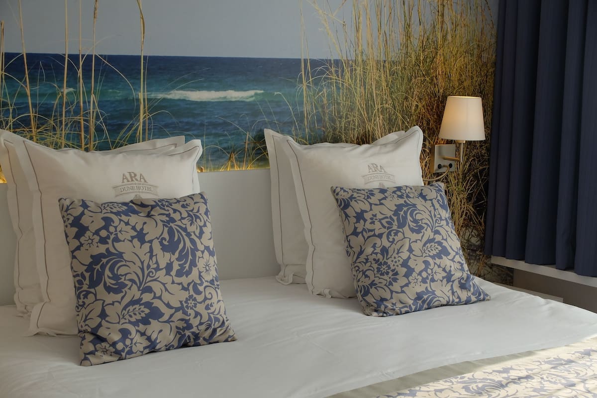 Uma cama com duas almofadas, uma luminária e vista para o oceano atrav és de grama alta.