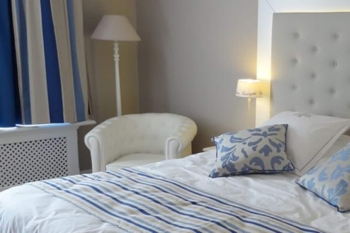 Quarto de hotel com uma cama grande, duas luminárias de cabeceira, uma poltrona branca e uma janela com cortinas.