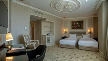 Quarto de hotel com duas camas, uma escrivaninha com luminária, uma cadeira, teto decorativo e um quadro na parede.