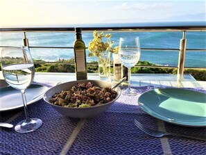 Mesa posta com uma saladeira, dois copos de vinho e uma garrafa, com vista para a paisagem costeira.