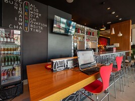 Uma área de bar com um laptop sobre uma mesa, uma geladeira com bebidas e uma parede com texto.