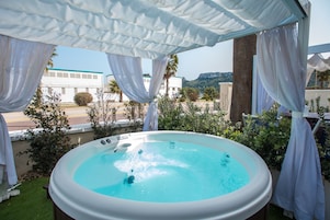 Uma banheira de hidromassagem sob um toldo branco com cortinas penduradas, rodeada por vegetação e com vista para edifícios e colinas.