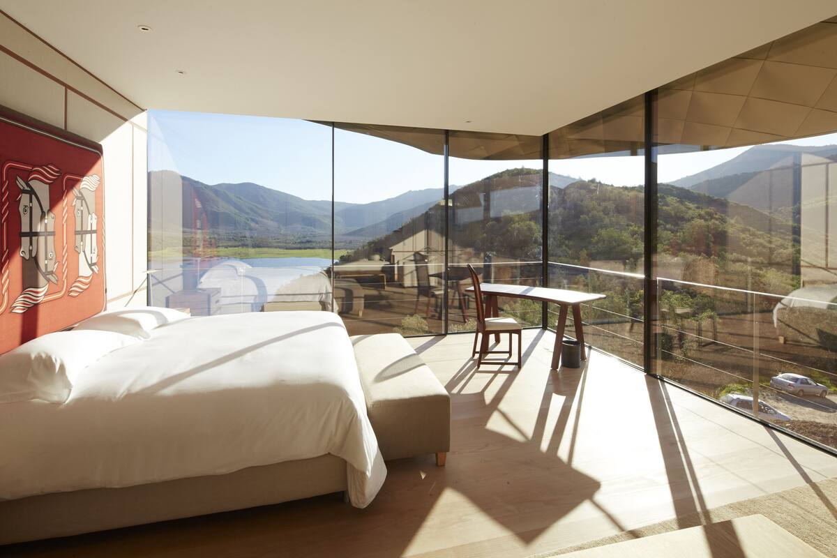 Quarto moderno com uma cama grande, uma mesinha e uma cadeira. Possui uma janela ampla com vista para as montanhas.