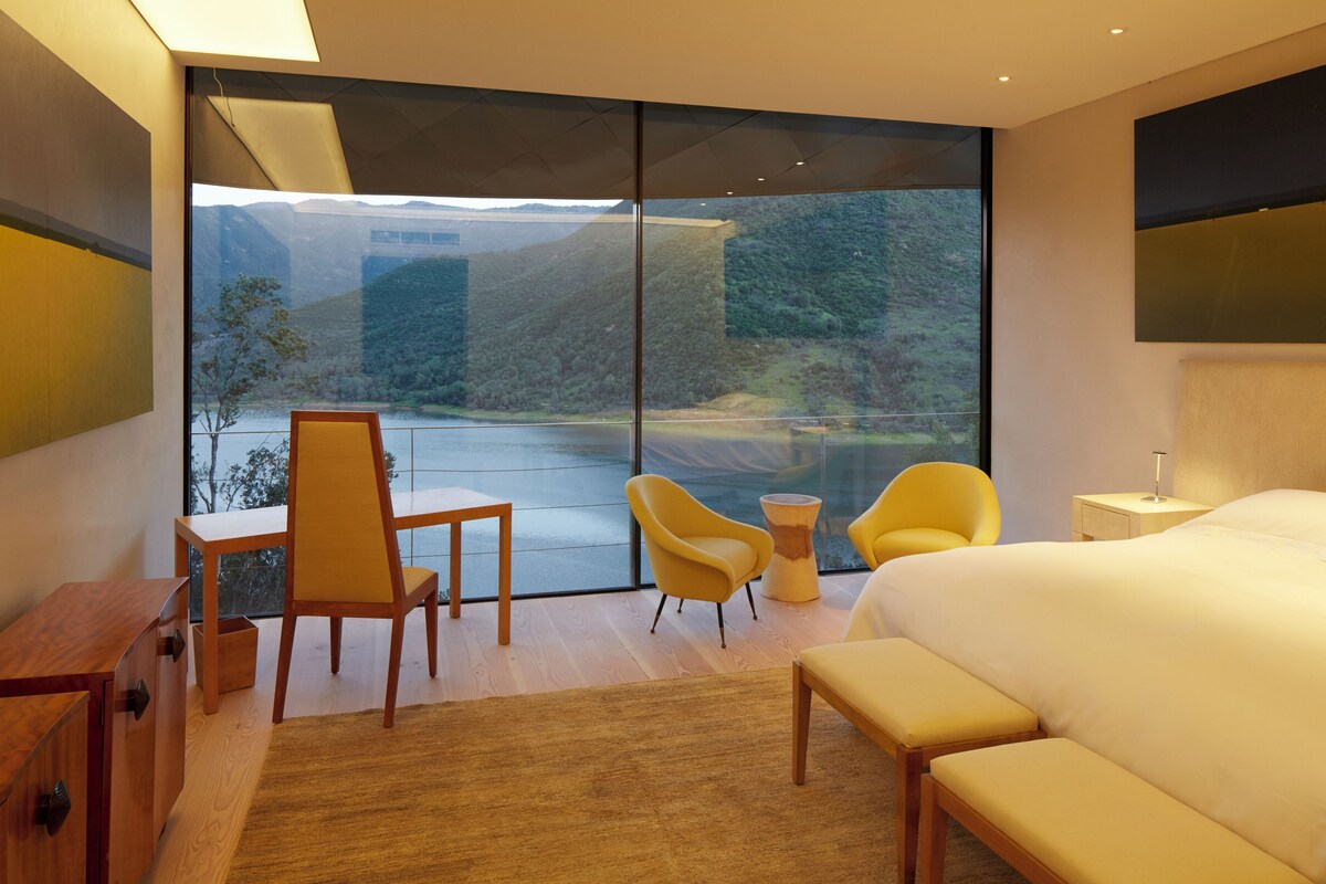 Quarto moderno com uma cama grande, uma escrivaninha, duas cadeiras e vista para um lago e montanhas.