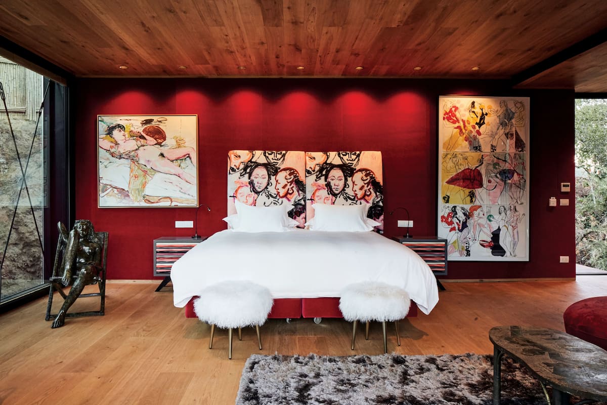 Quarto moderno com teto de madeira, cama grande com roupa de cama branca e três grandes pinturas nas paredes.