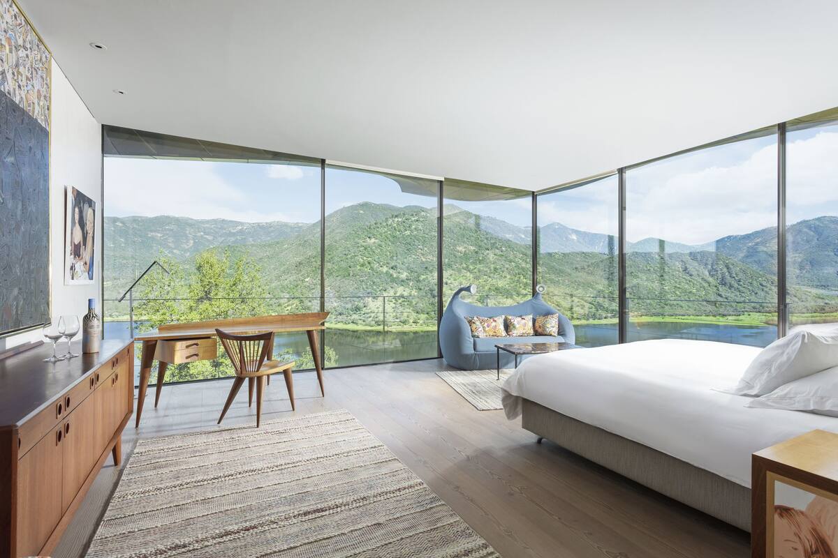 Quarto com uma cama grande, uma escrivaninha e uma cadeira, com vista para montanhas e um lago.