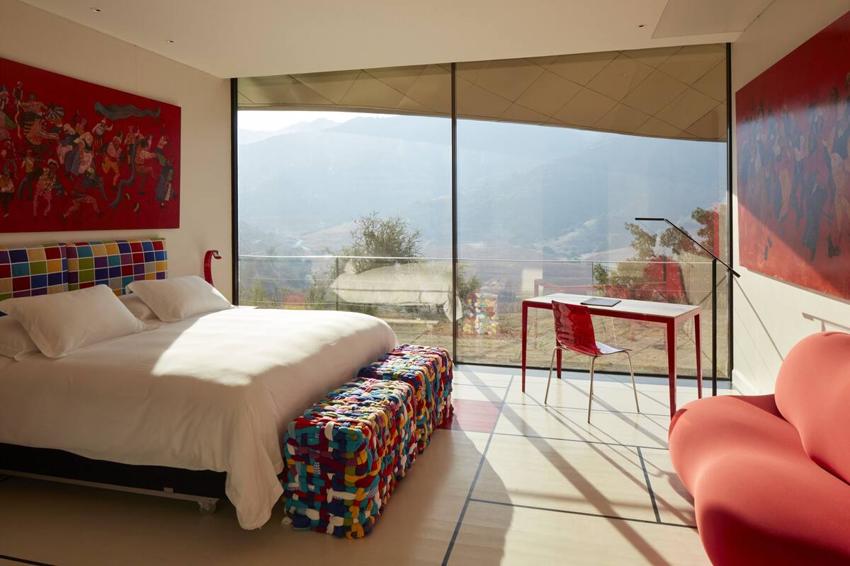 Quarto moderno com cama grande, um banquinho acolchoado colorido, uma cadeira vermelha e uma parede de vidro com vista para as montanhas.