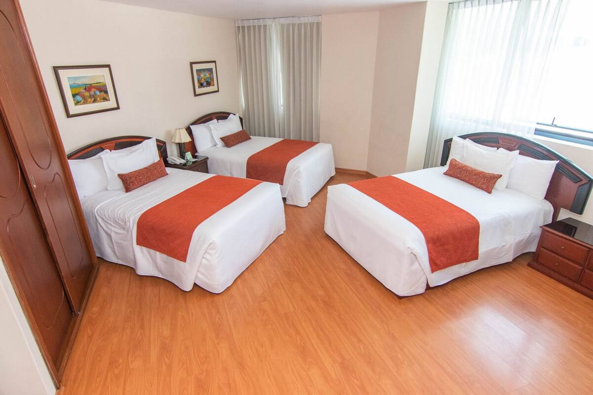 Quarto de hotel com duas camas, piso de madeira, mesa de cabeceira com luminária e duas imagens emolduradas na parede.