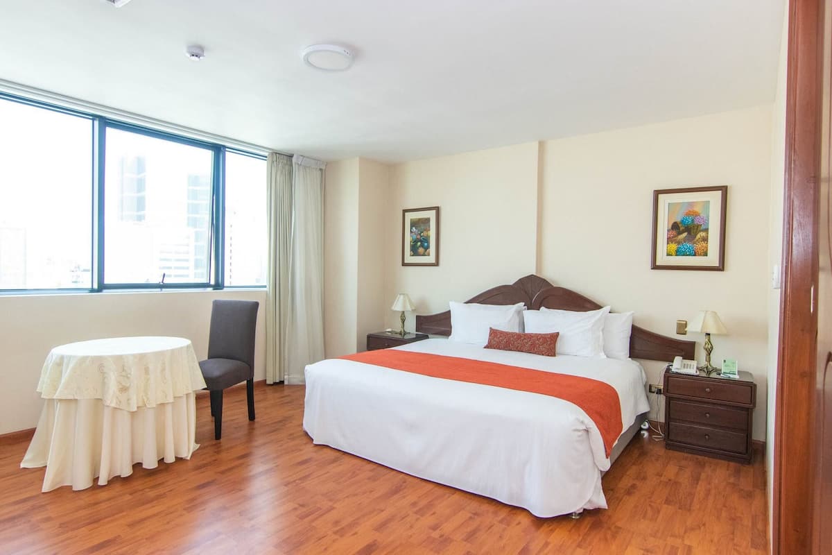 Quarto de hotel com uma cama grande, uma mesa redonda e duas mesinhas de cabeceira.