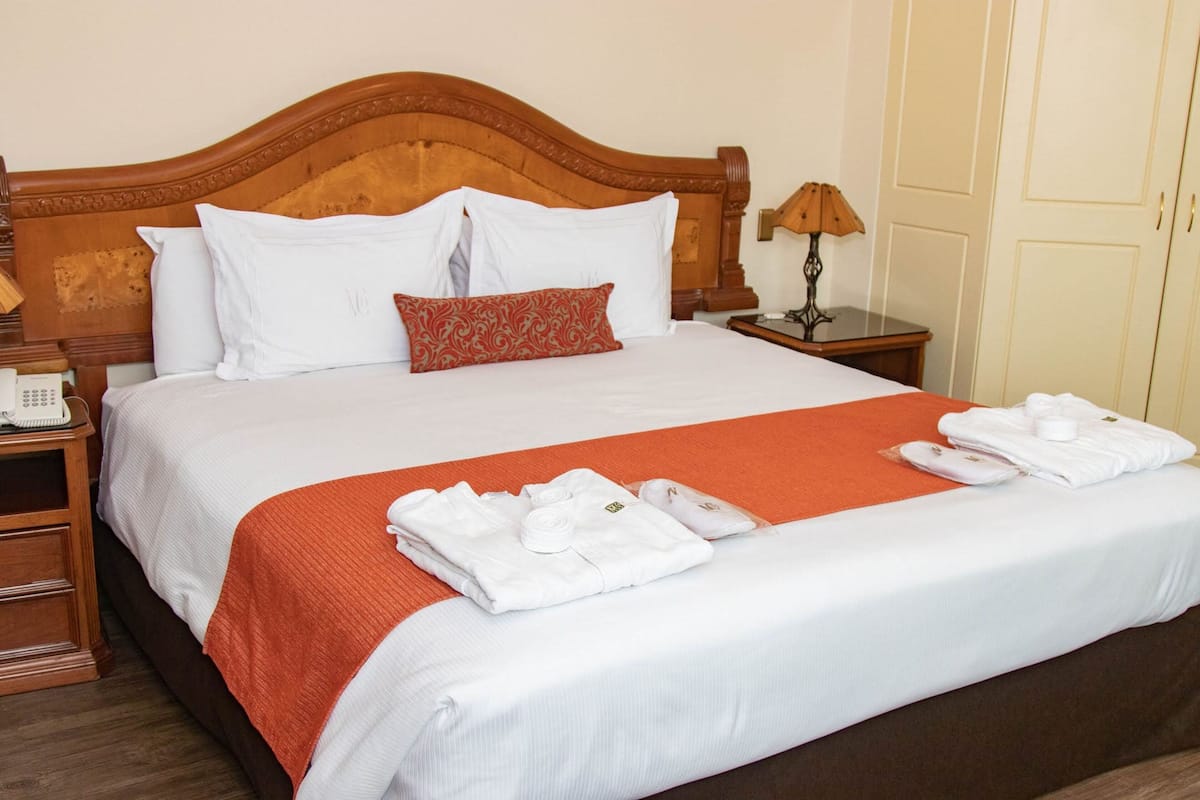 Uma cama bem arrumada com roupa de cama branca e vermelha, uma cabeceira de madeira e uma mesa de cabeceira com abajur e telefone.