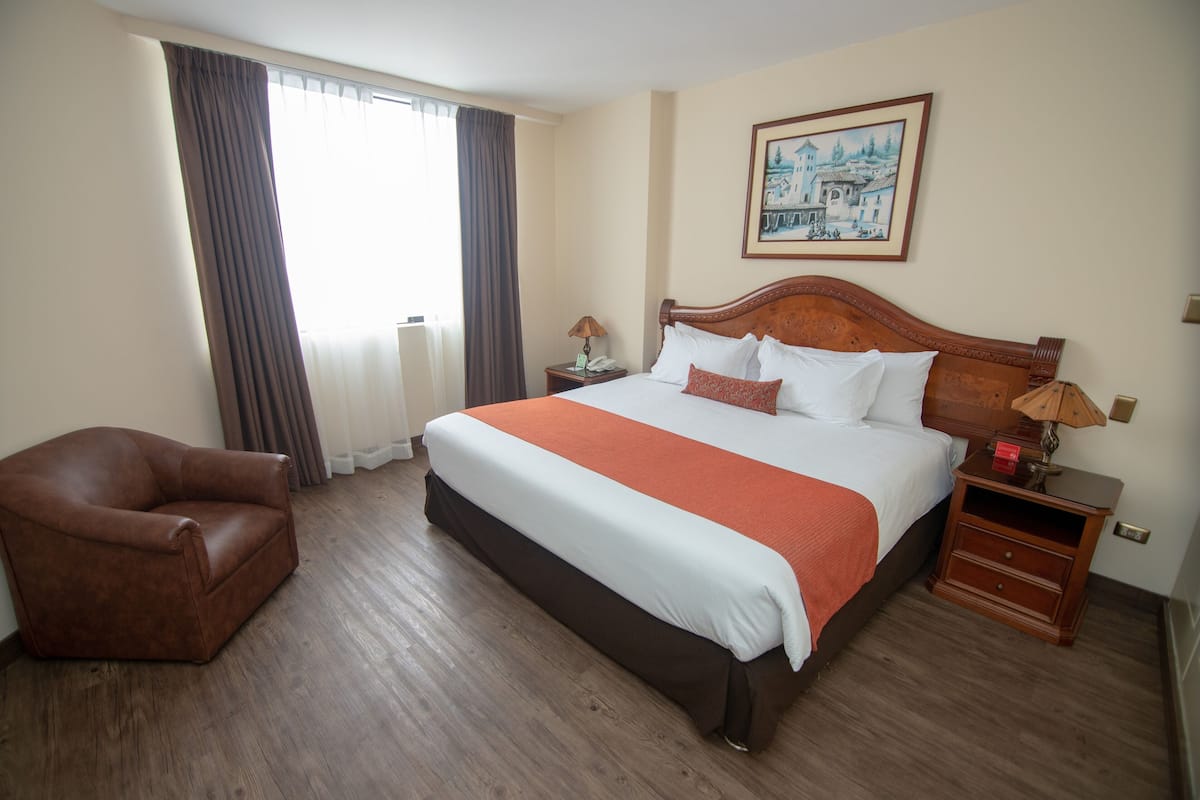 Quarto de hotel com uma cama grande, uma mesa de cabeceira de madeira, uma poltrona de couro marrom e uma janela com cortinas.