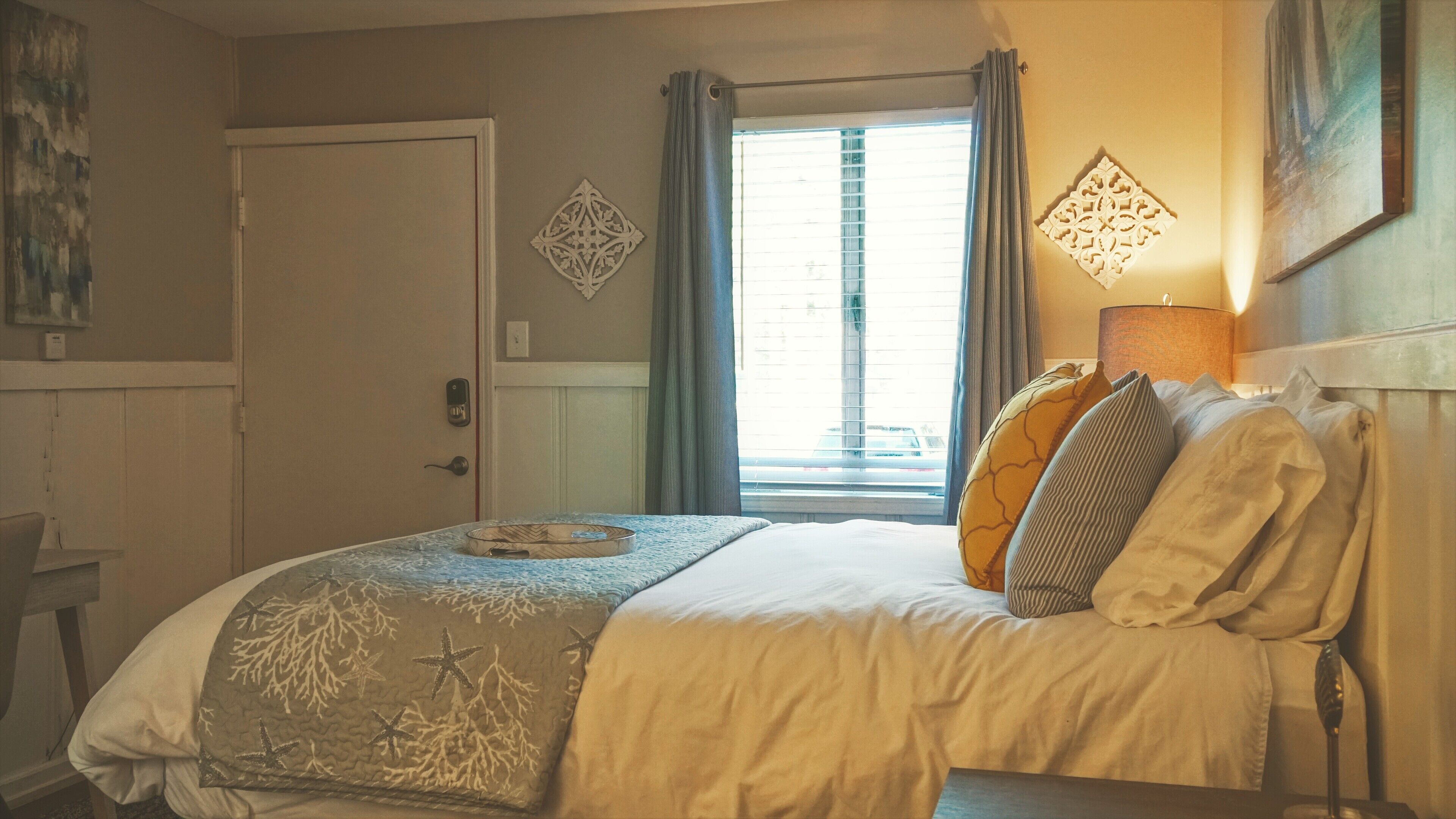 Um quarto com cama, mesa de cabeceira, luminária, janela com cortinas e porta.