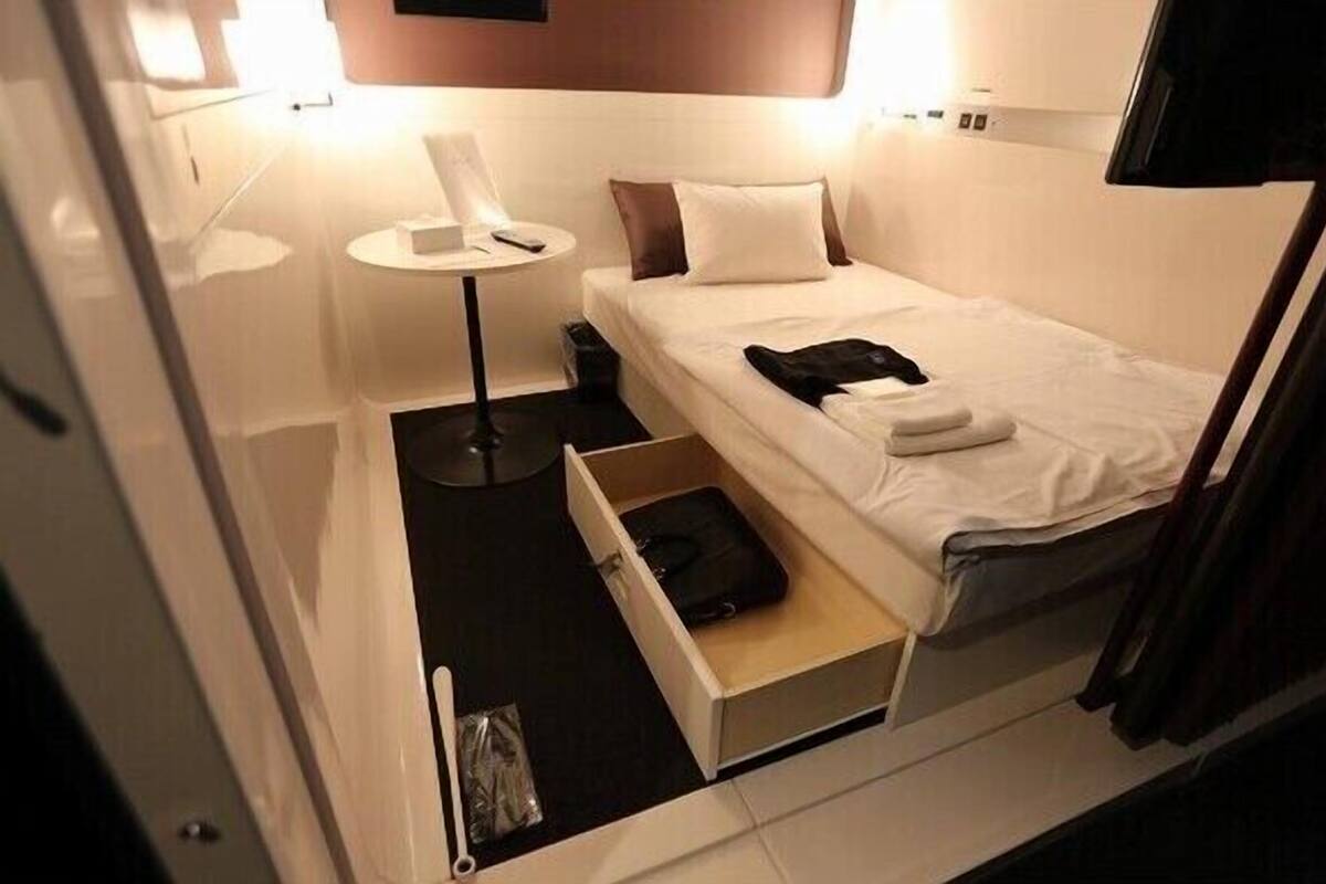Quarto de hotel pequeno com uma cama de solteiro, mesa de cabeceira, escrivaninha e gaveta para guardar objetos.