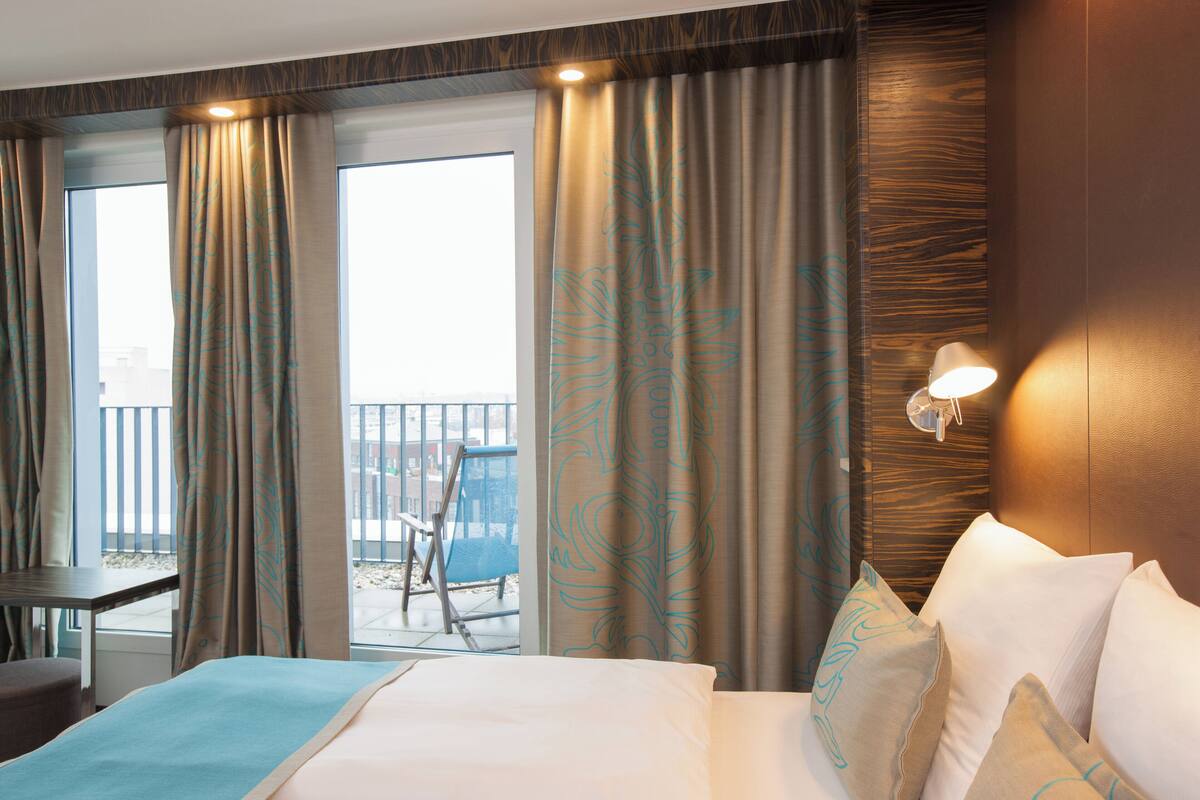 Quarto de hotel com cama, cortinas, mesa de cabeceira e vista para a varanda externa.