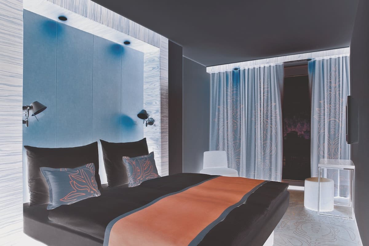 Quarto moderno com uma cama grande, cabeceira com iluminação embutida, mesa de cabeceira e vista para o exterior através de cortinas.