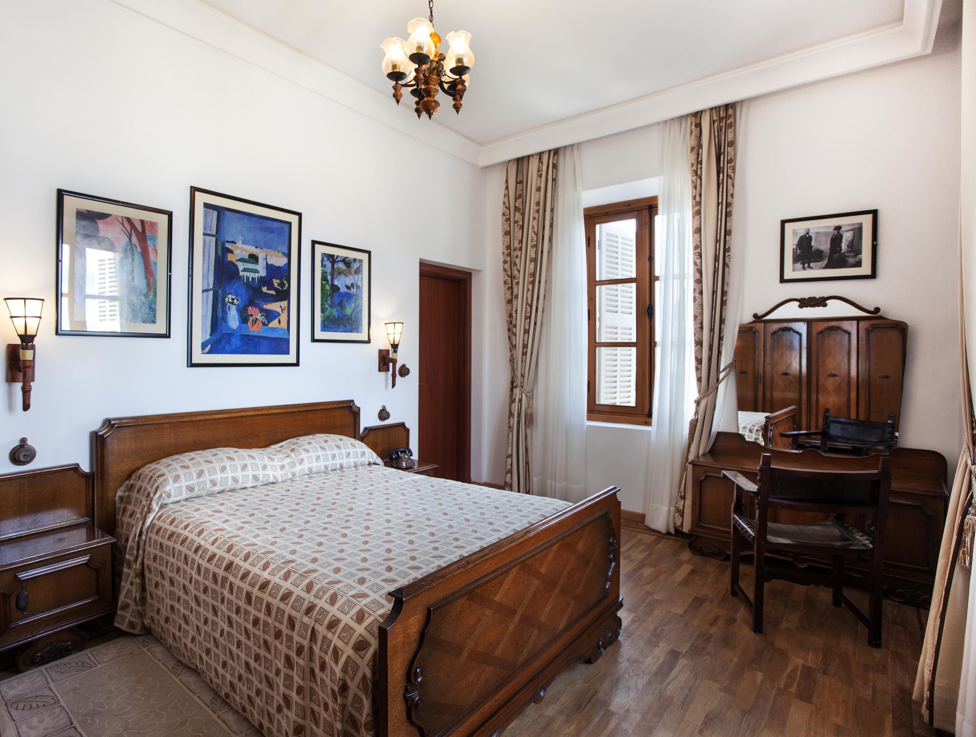 Quarto com cama de madeira, escrivaninha e cadeira. Há três quadros na parede, um lustre e uma janela com cortinas.