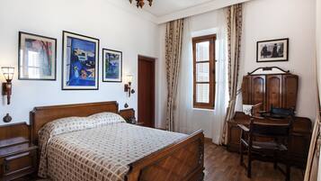 Quarto com cama de madeira, escrivaninha e cadeira. Há três quadros na parede, um lustre e uma janela com cortinas.