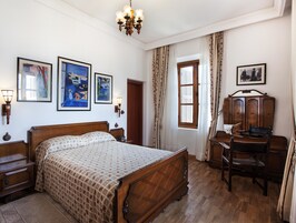 Quarto com cama de madeira, escrivaninha e cadeira. Há três quadros na parede, um lustre e uma janela com cortinas.