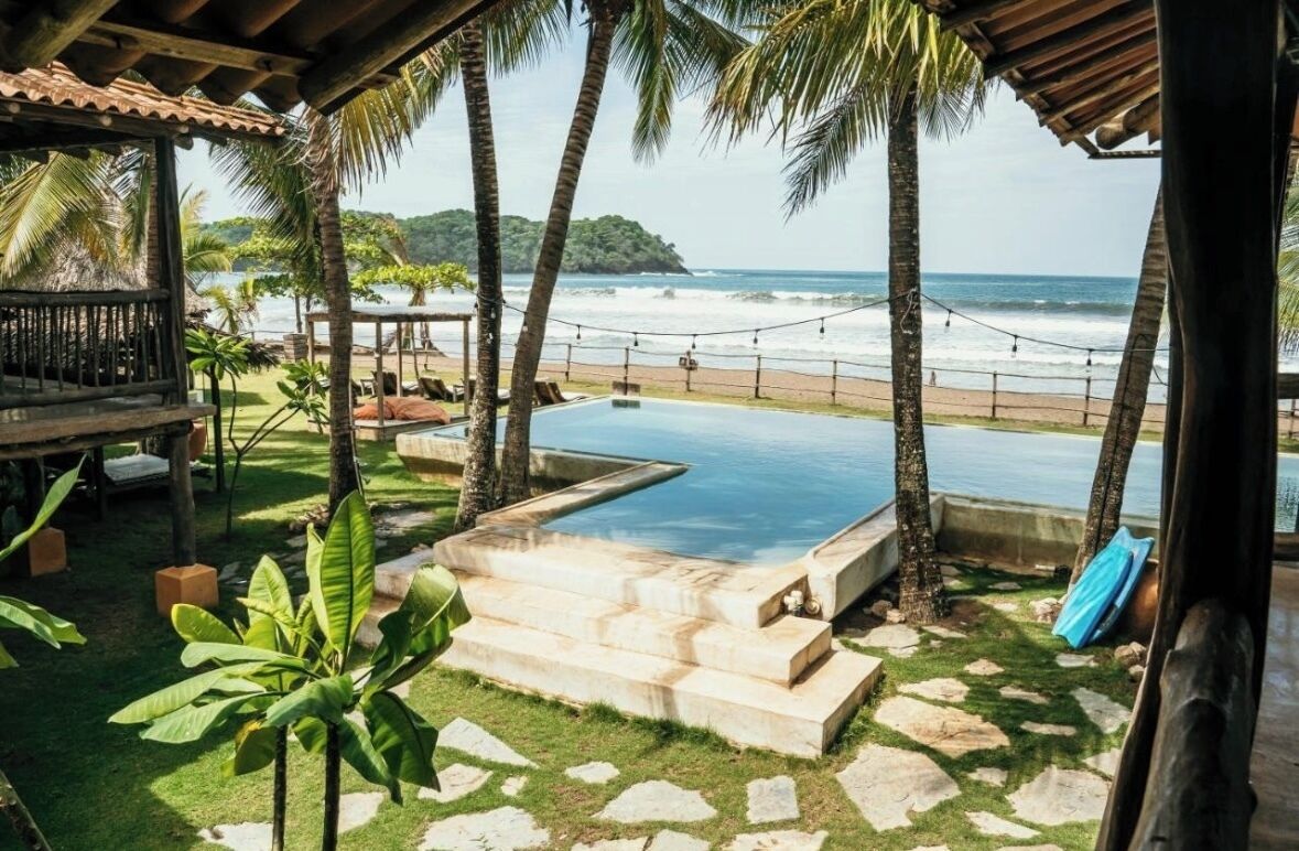Piscina infinita com vista para o oceano, cercada por palmeiras e um deck de madeira.