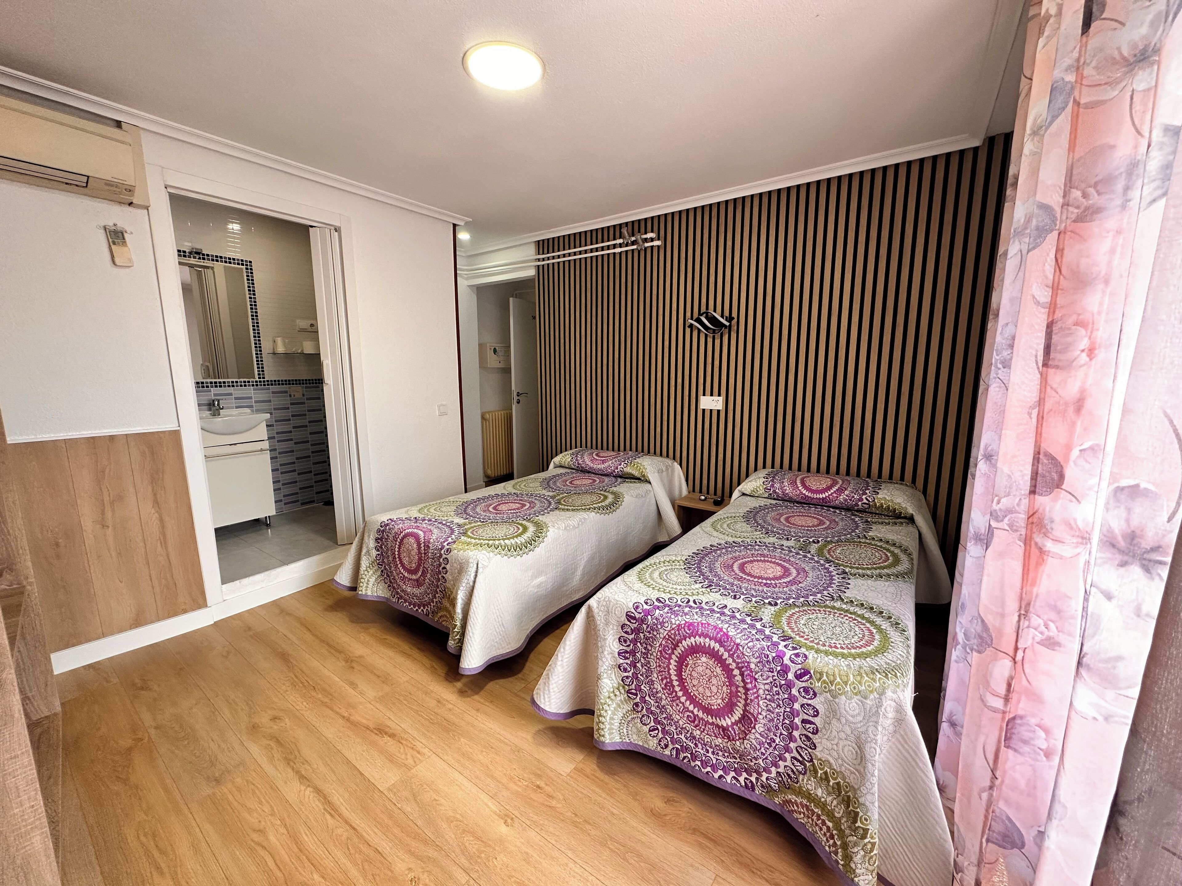 Quarto com duas camas, piso de madeira, banheiro com chuveiro e parede listrada.