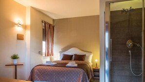 Quarto de hotel com cama, mesa de cabeceira com planta, área de chuveiro e janela com cortinas.