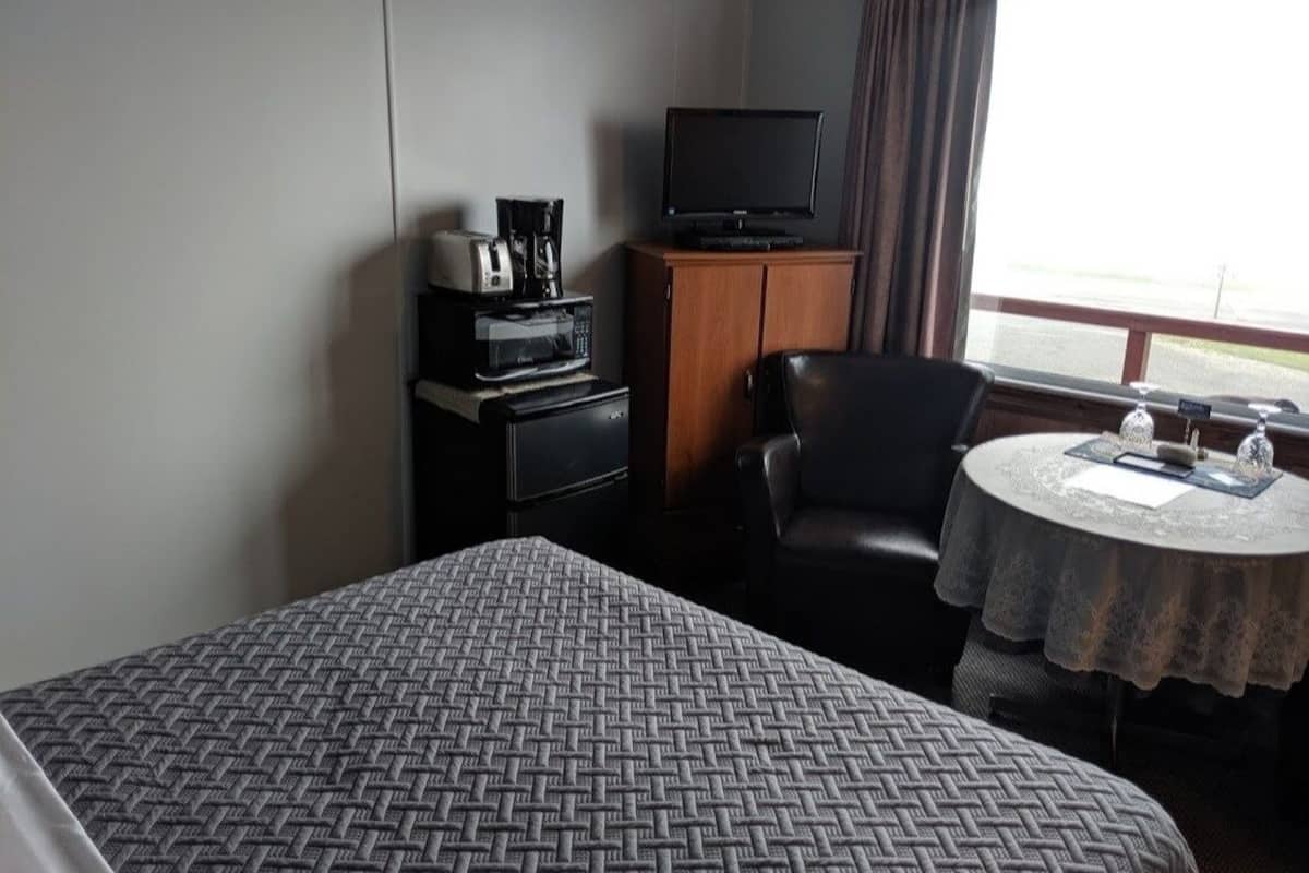 Quarto de hotel com cama, televisão sobre um móvel de madeira, cafeteira, poltrona e mesa com toalha de renda.