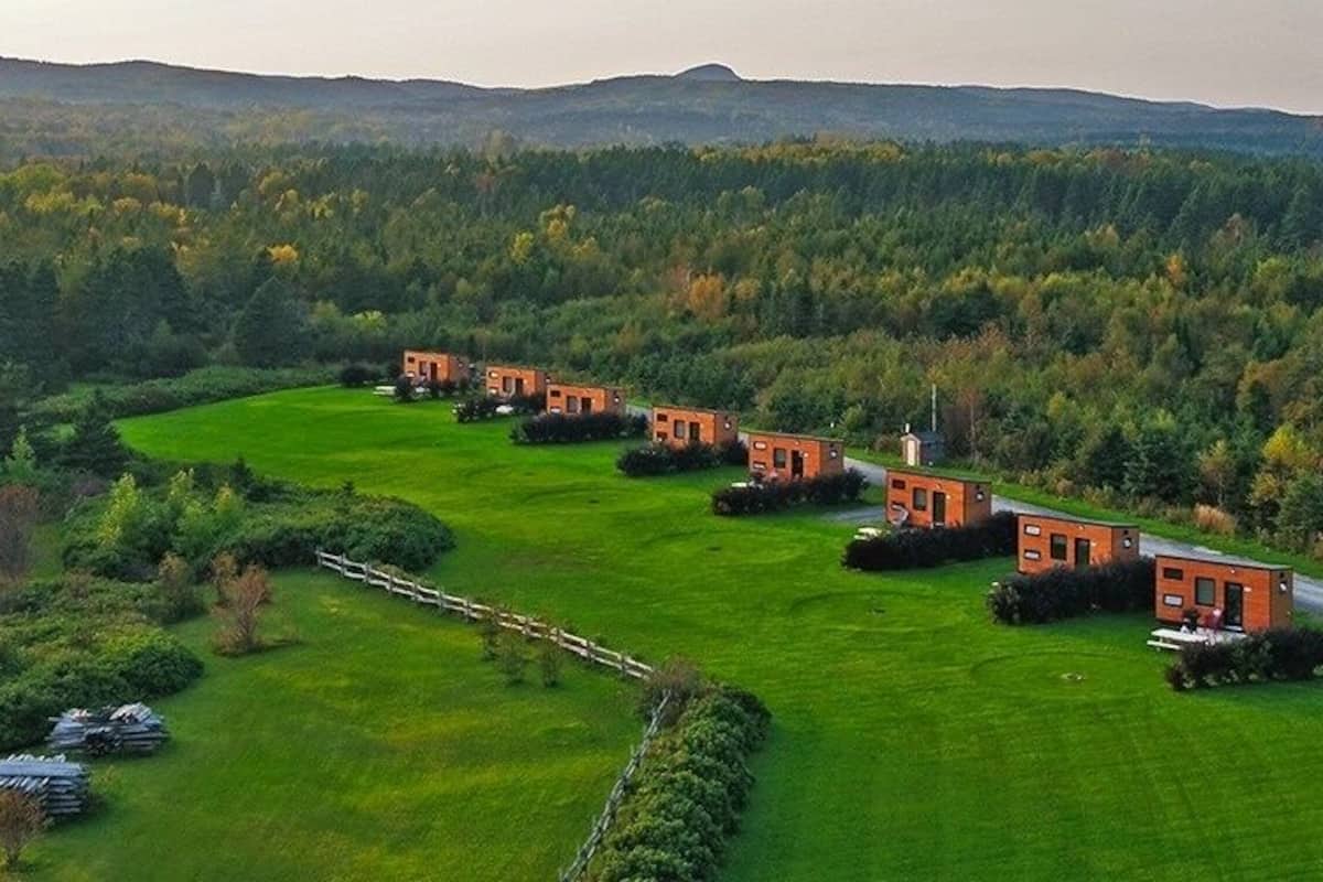 Uma série de pequenas cabanas rústicas dispostas em semicírculo sobre um campo verdejante, rodeadas por uma floresta densa.
