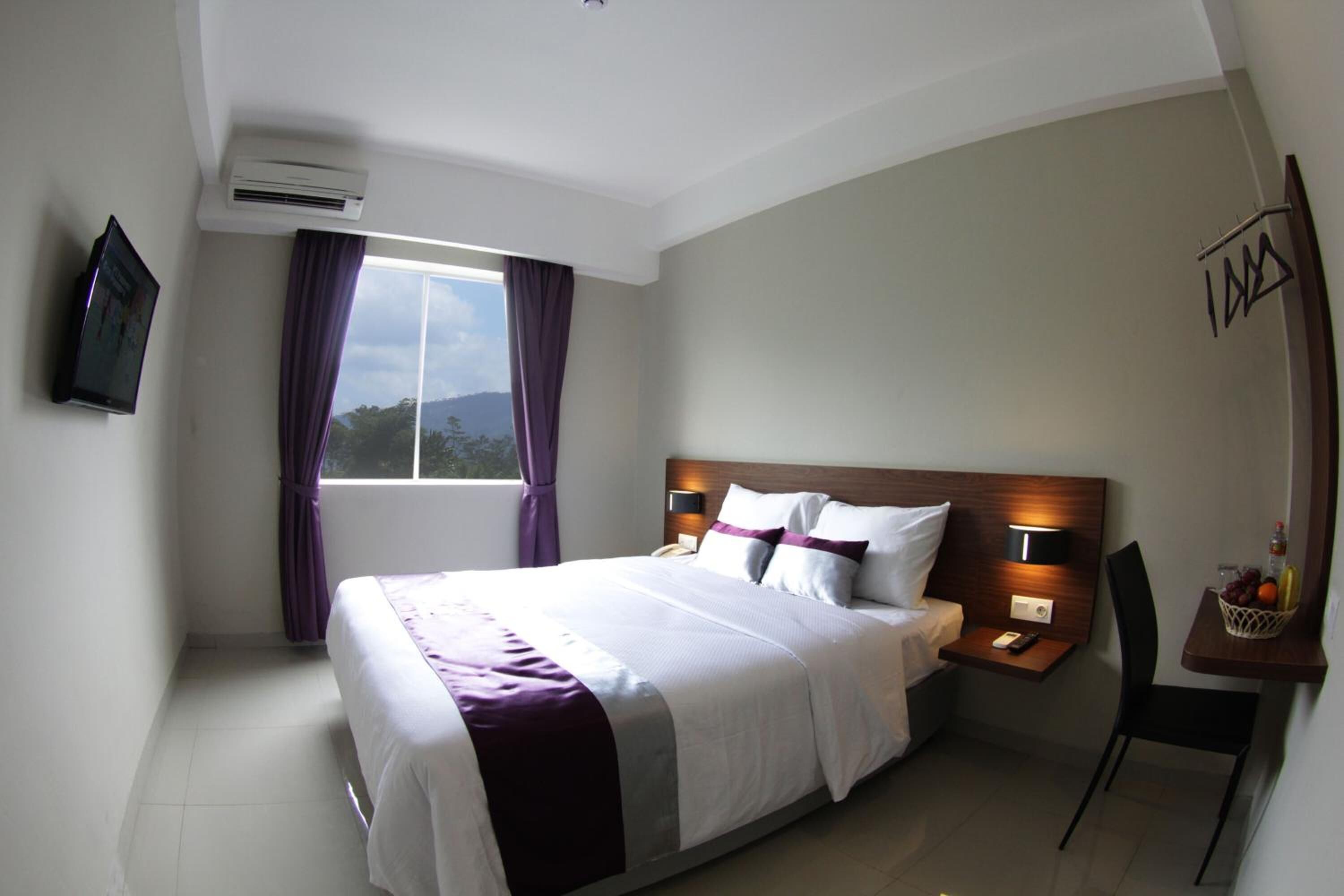 Quarto de hotel com cama, mesa de cabeceira, escrivaninha e cadeira, televisão na parede e janela com cortinas.