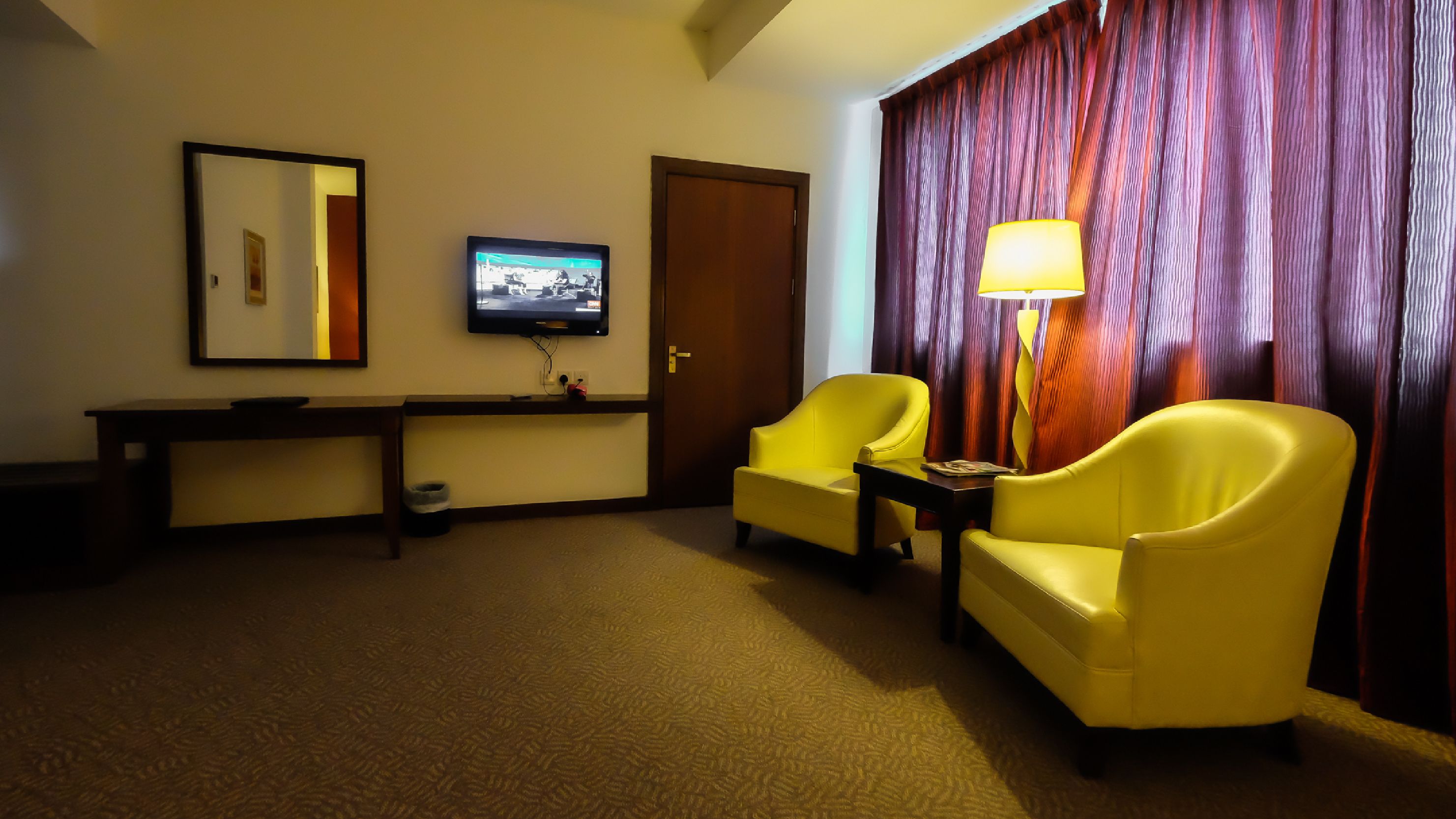 Quarto de hotel com televisão, escrivaninha, duas poltronas amarelas, luminária e uma porta.