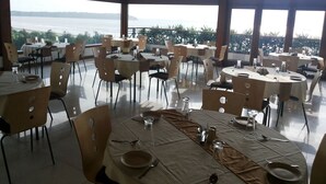 Um restaurante com várias mesas e cadeiras, janelas amplas com vista para o mar e teto alto.