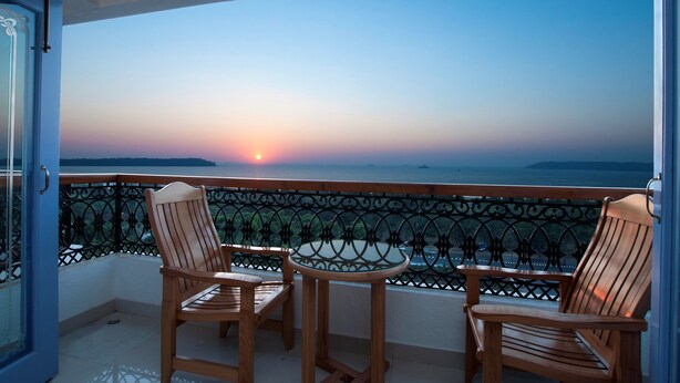 Varanda com duas cadeiras de madeira e uma mesa redonda pequena, com vista para um pôr do sol sobre a água e terraços ao longe.