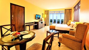 Quarto de hotel com cama, sofá, mesa de jantar com frutas, televisão e janela com cortinas.