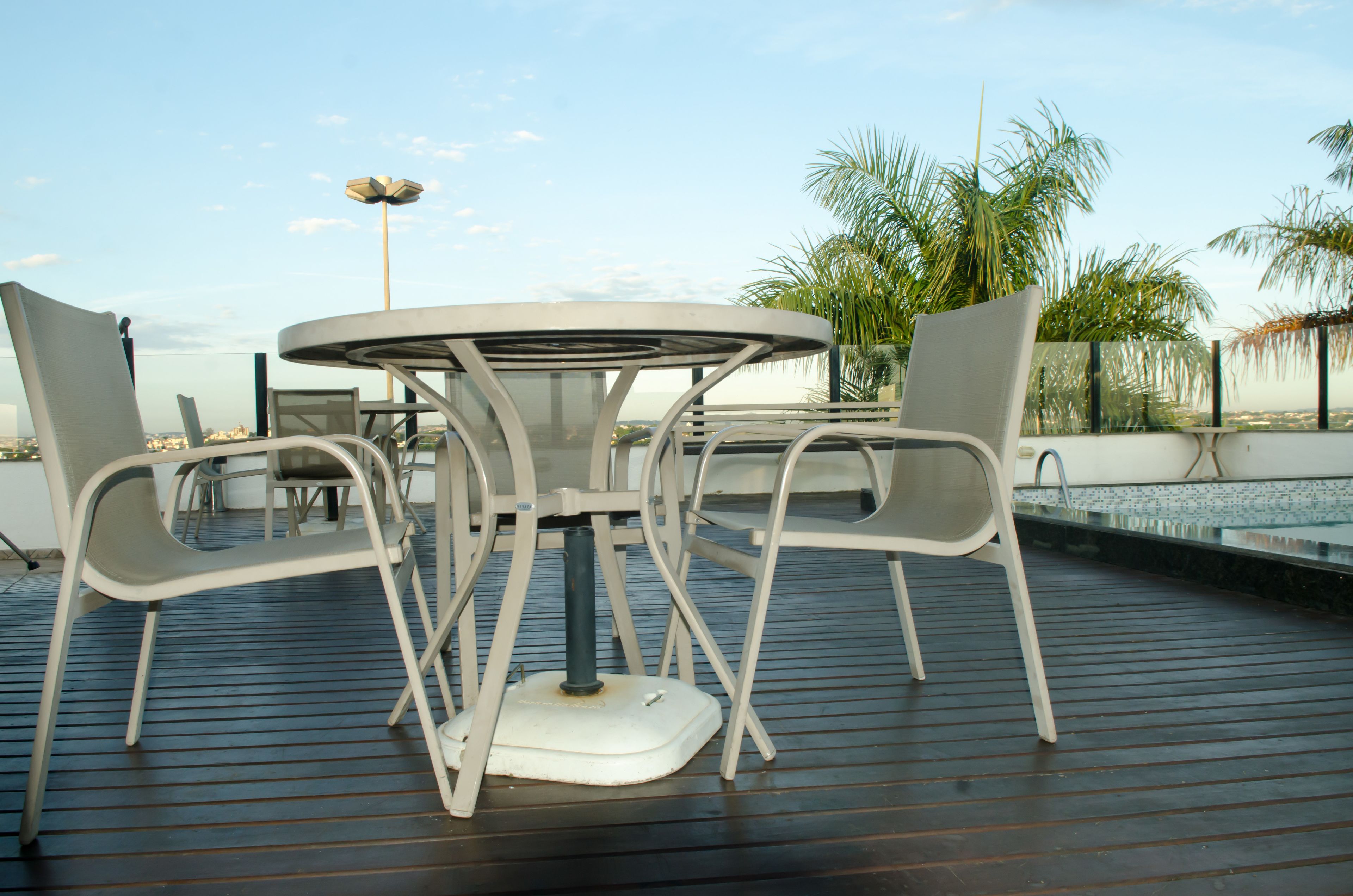 Mesa redonda com quatro cadeiras em deck de madeira, com vista para área de piscina e palmeiras.