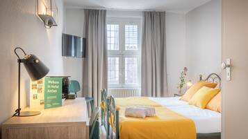 Quarto de hotel com uma cama, uma escrivaninha com abajur, televisão e janela com cortinas.