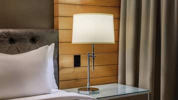 Uma cama bem arrumada com travesseiro branco, uma mesa de apoio de vidro com abajur e lambris na parede.