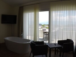 Quarto de hotel com banheira, TV fixada na parede, varanda com mesa e cadeiras e vista para a cidade e o mar.