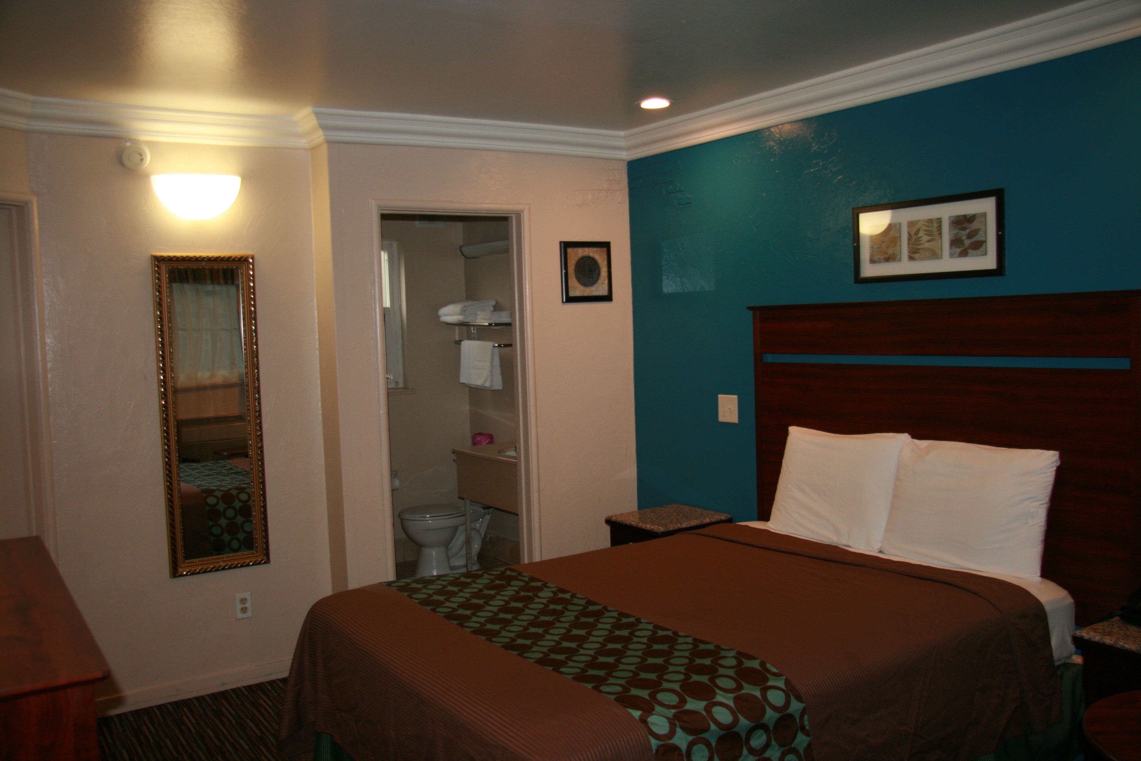 Quarto de hotel com uma cama, mesa de cabeceira, espelho, banheiro visível por uma porta aberta e um quadro na parede.
