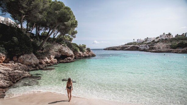 Uma praia com águas turquesa cristalinas, areal arenoso e falésias rochosas com vegetação.
