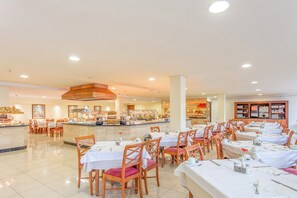 Uma ampla área de refeições com várias mesas e cadeiras, um buffet central e um teto bem iluminado com iluminação embutida.