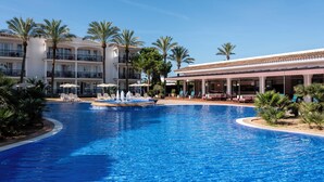 Uma piscina grande com espreguiçadeiras e guarda-sóis, cercada por palmeiras e um edifício de v ários andares com varandas.