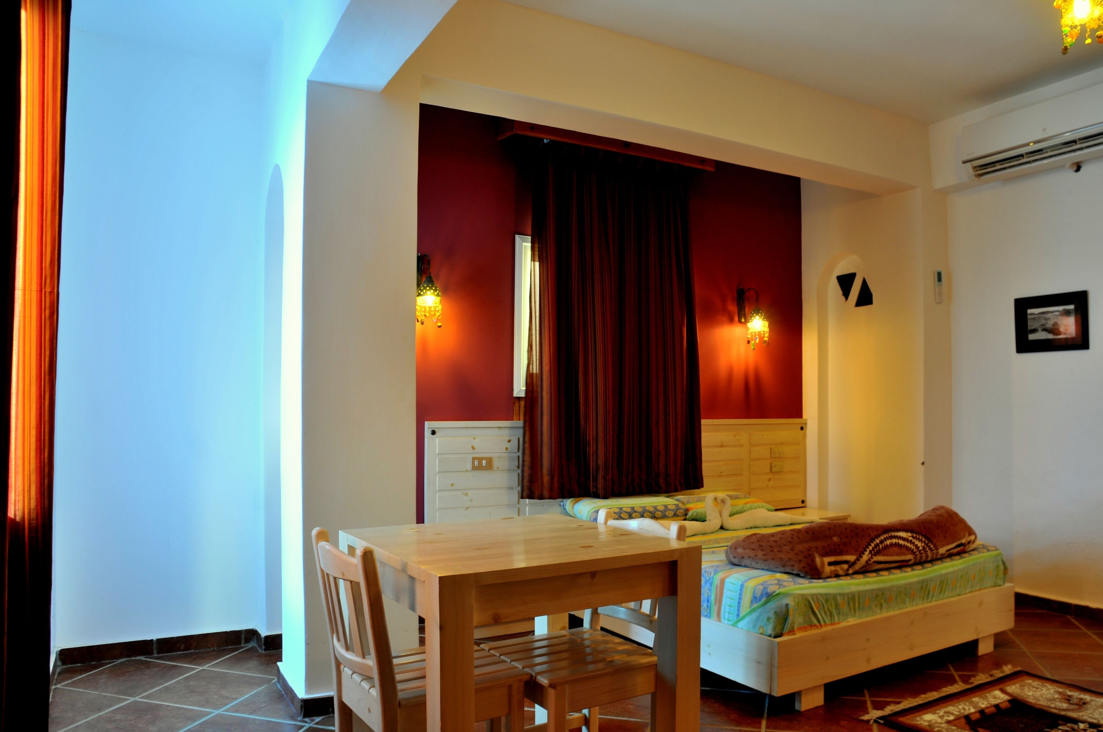 Quarto com uma cama, uma mesa de madeira e uma cadeira. Há uma janela com cortinas, um ar-condicionado fixado na parede e um quadro na parede.