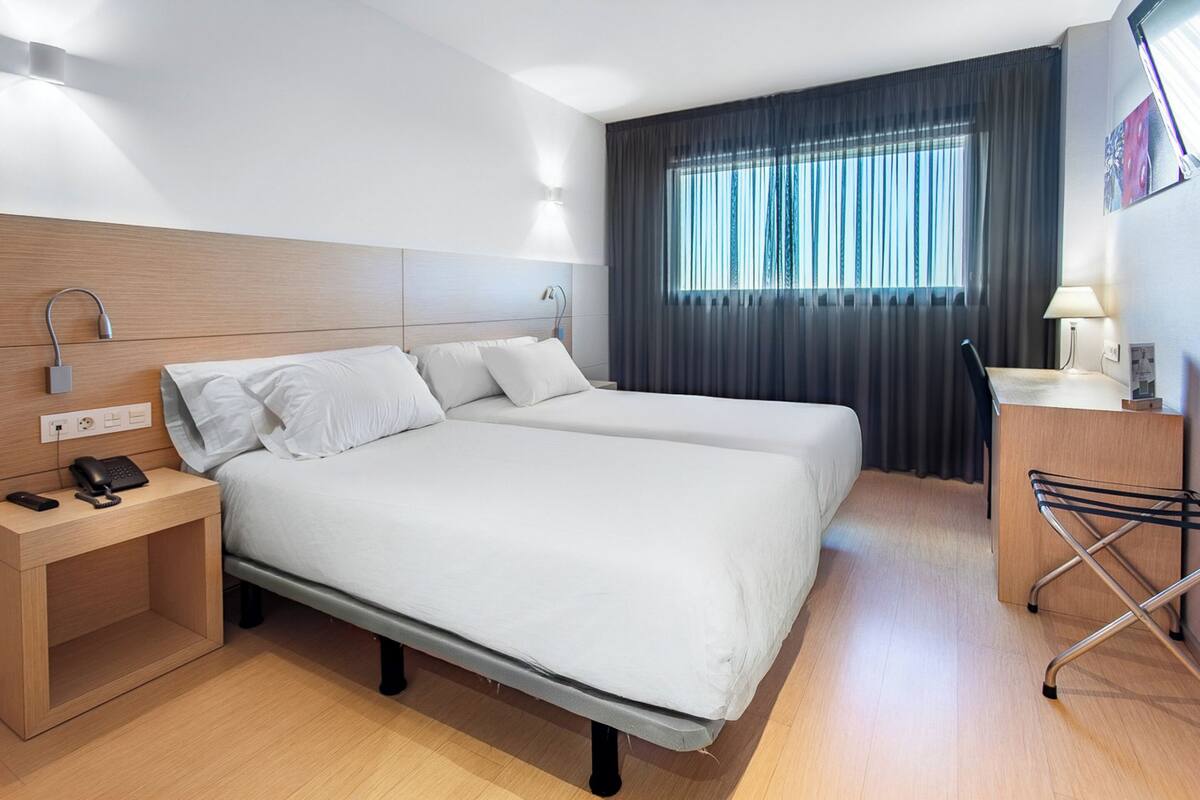 Quarto de hotel com cama, mesa de cabeceira com telefone, escrivaninha com abajur e janela com cortinas.