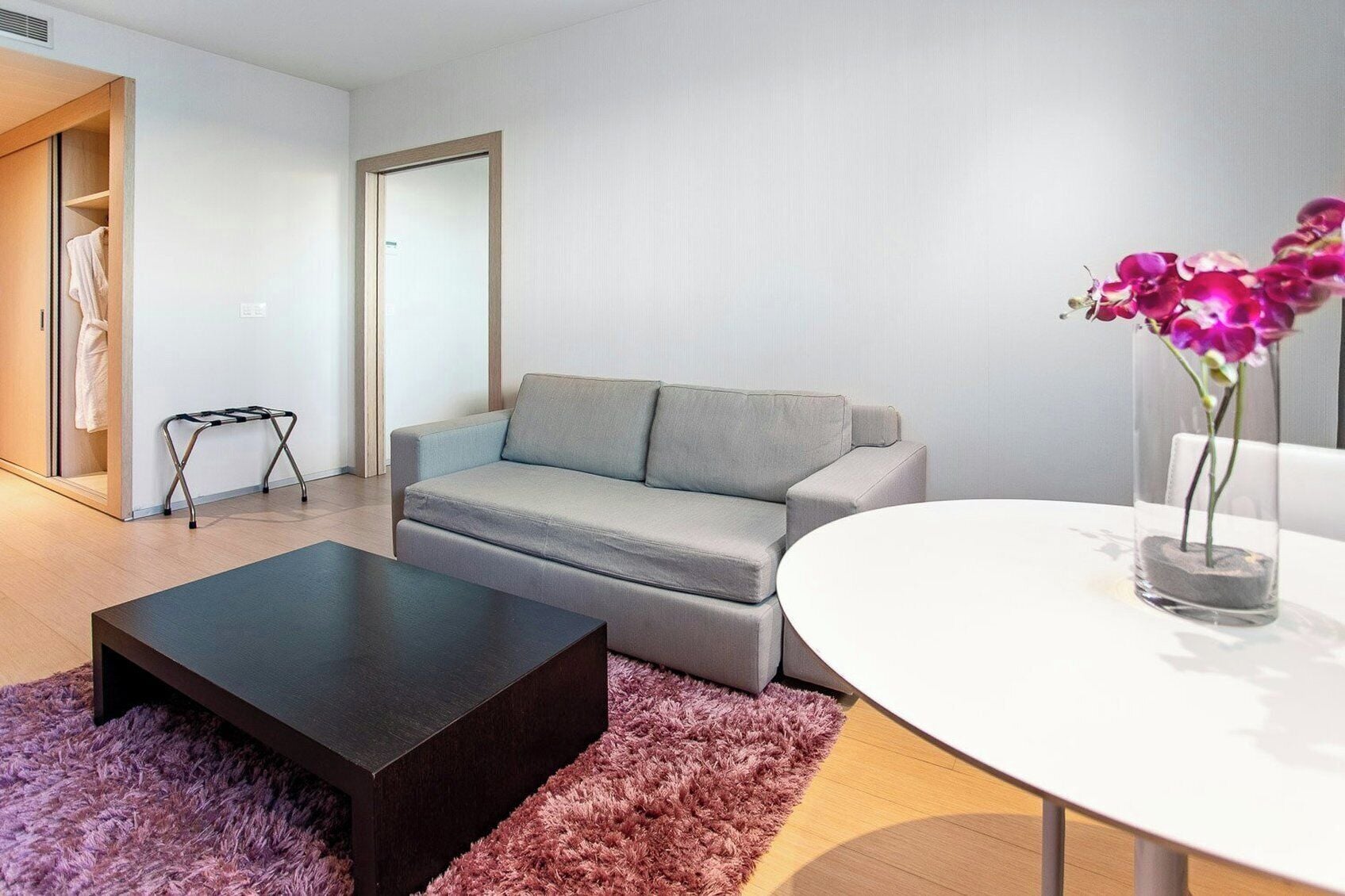 Uma sala de estar moderna com um sofá cinzento, uma mesa de centro escura, uma mesa redonda branca com um vaso de flores, um tapete roxo e um banco de madeira.