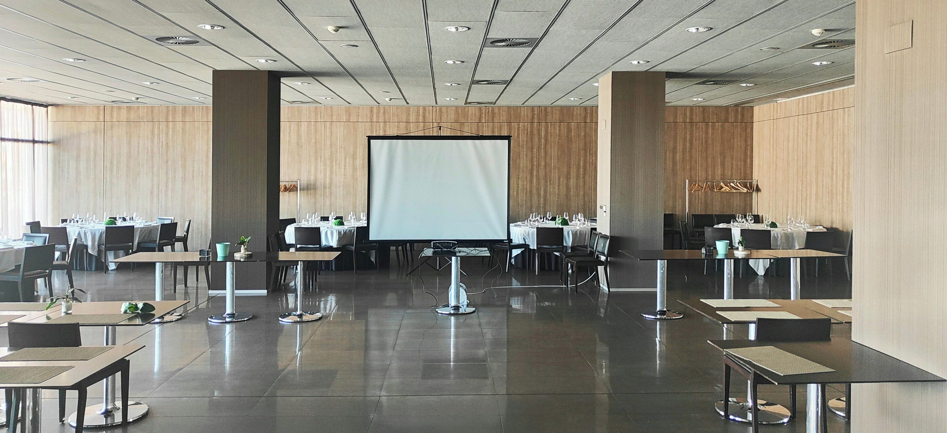 Uma sala de conferência com mesas, cadeiras, uma tela de projetor e uma parede de madeira.