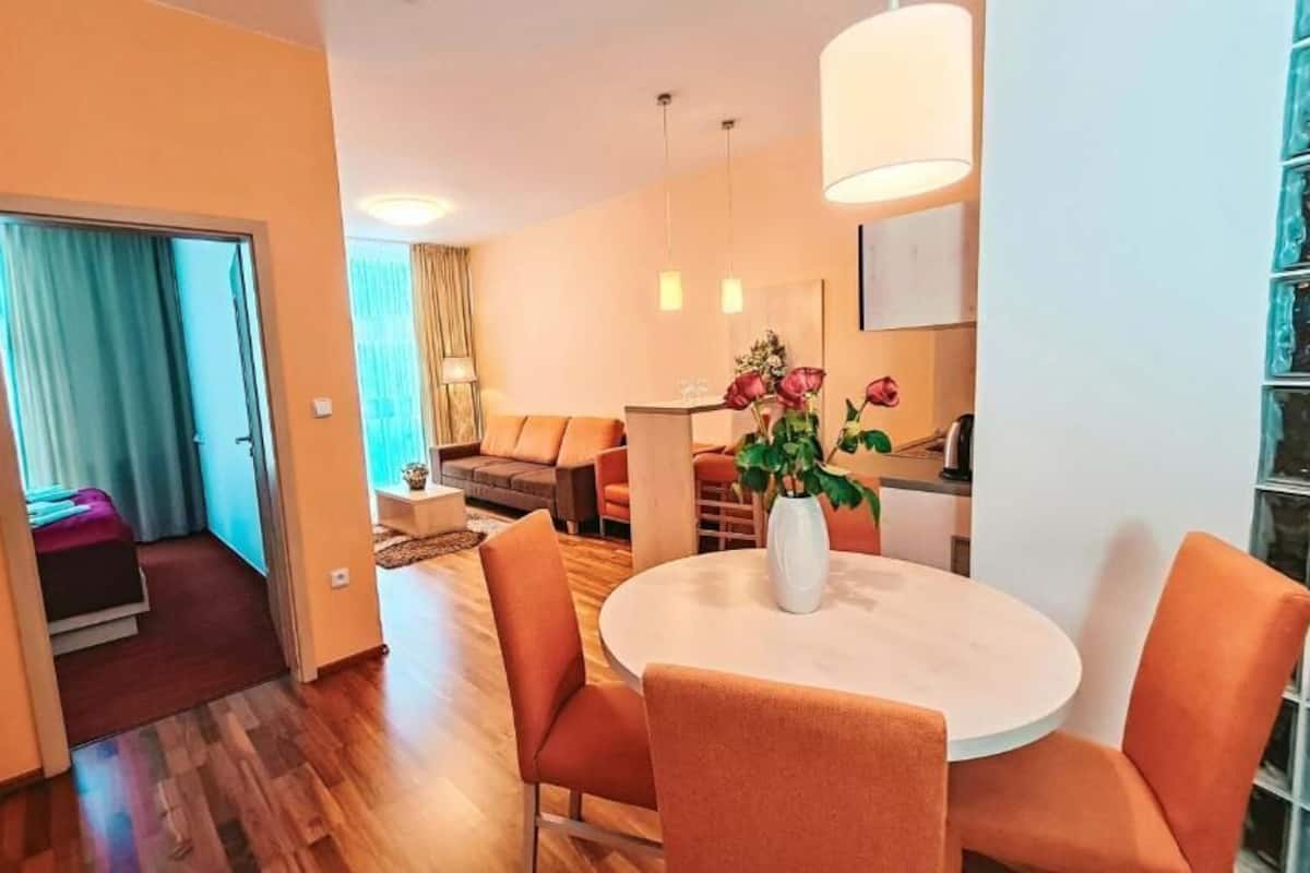 Uma sala de estar moderna com uma mesa de jantar redonda, cadeiras laranja, um sofá e uma janela grande com vista.