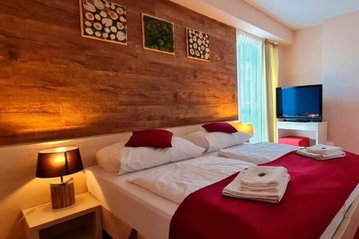 Quarto de hotel com cabeceira de madeira, cama com lençóis brancos e almofadas vermelhas, TV sobre um suporte e um tapete vermelho.