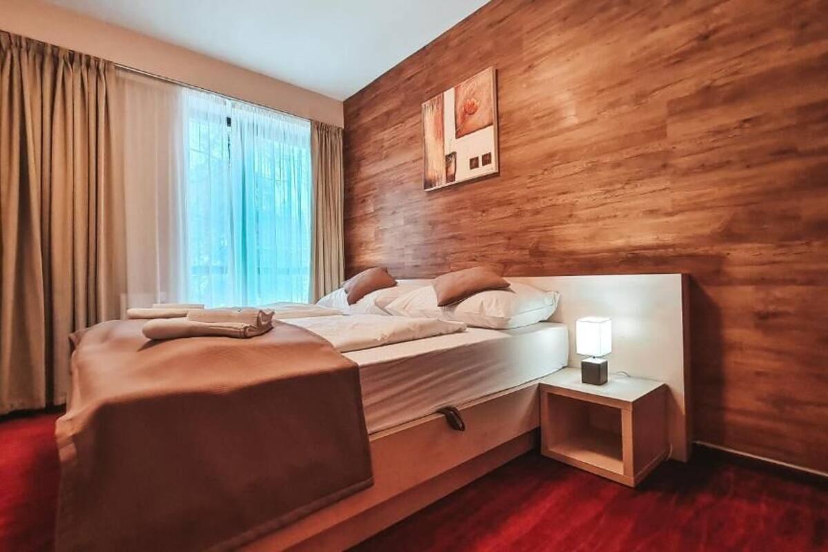 Quarto de hotel moderno com parede de madeira, uma cama grande com cabeceira, uma mesa de cabeceira com abajur e uma janela com cortinas.